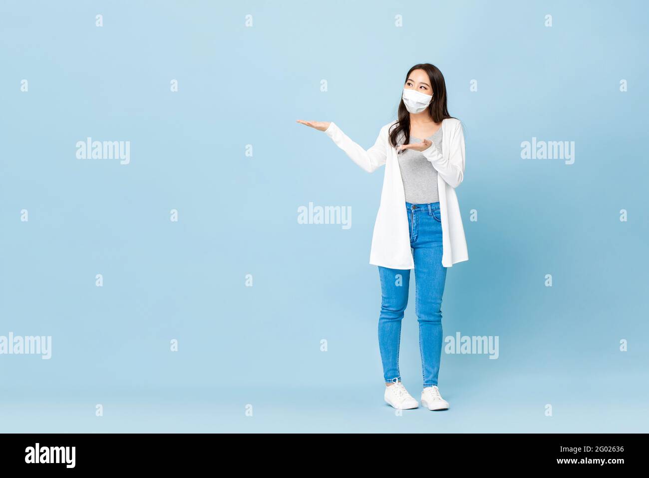 Young Asian woman wearing face mask opening hands to empty space aside ...