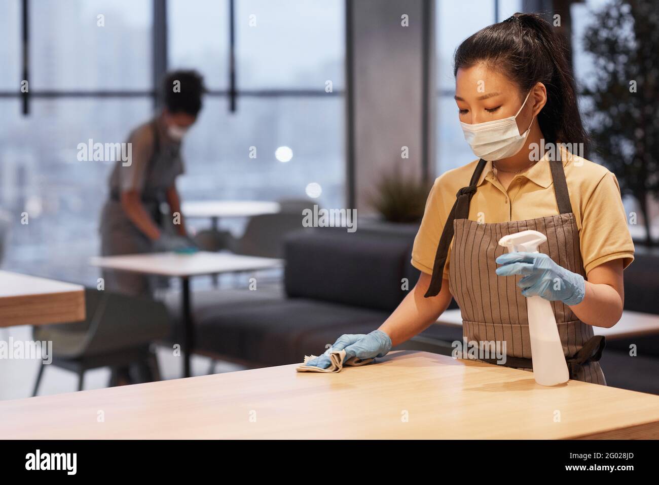 Portrait of young woman wearing mask while cleaning tables in modern ...
