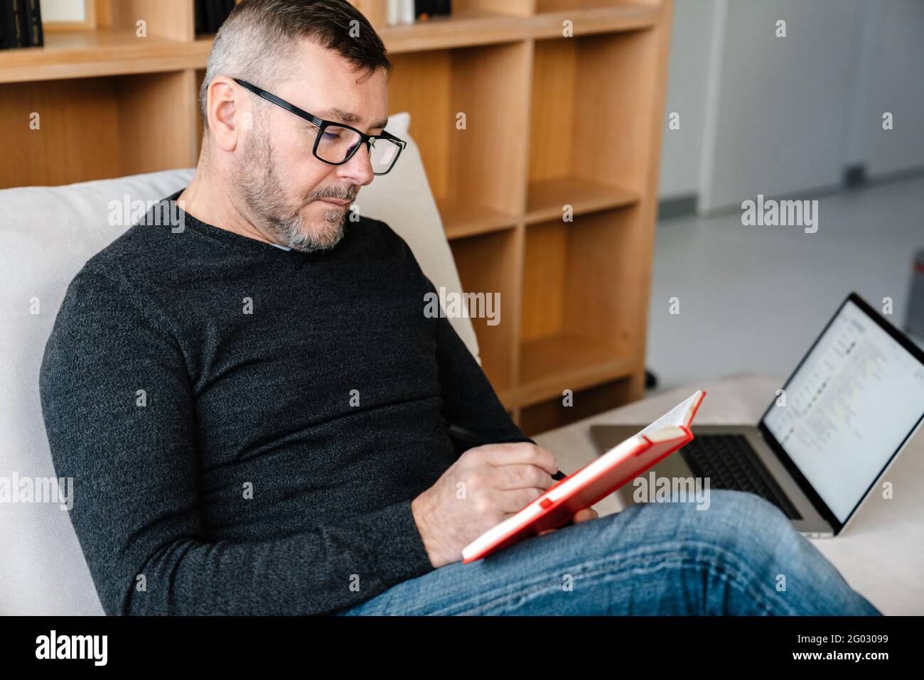 Serious adult unshaven man writing down notes while working with laptop ...