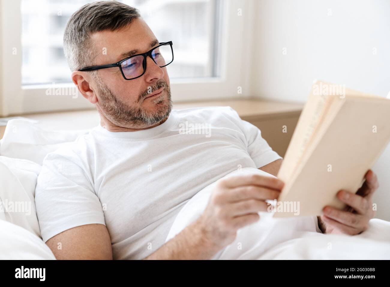 Serious adult unshaven man reading book while lying in bed at home ...