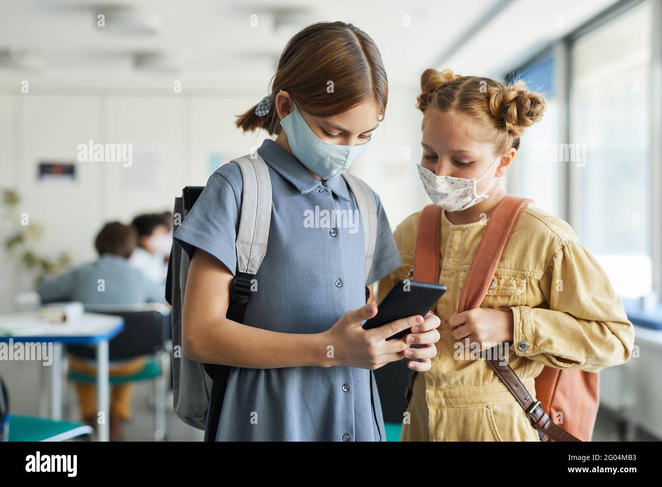 Waist up portrait of two girls wearing masks in school classroom and ...