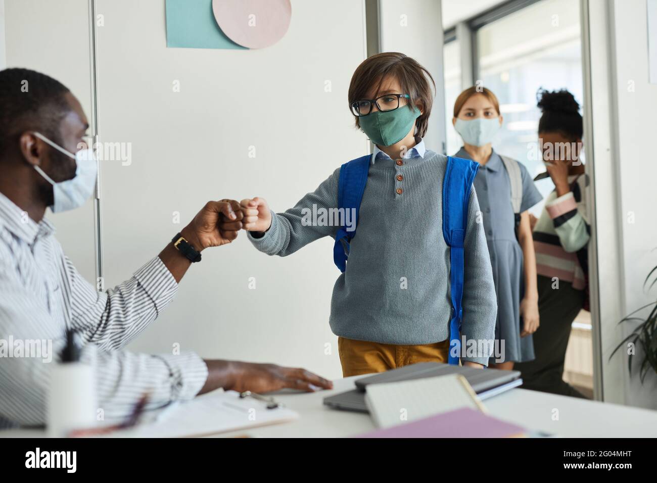 Portrait of male teacher wearing mask and fist bumping kids entering ...