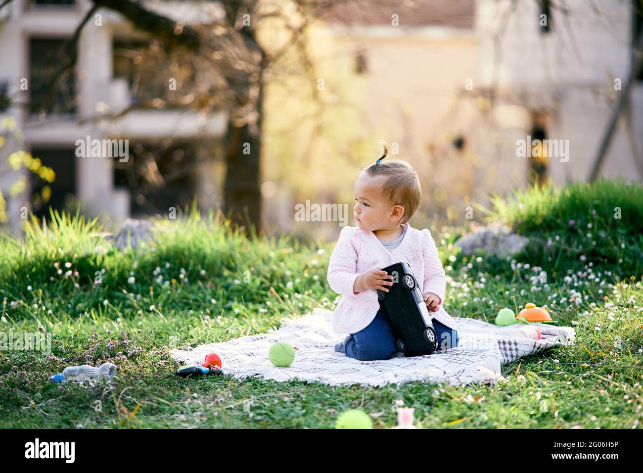 Little girl sits on a blanket among toys on green grass and holds a toy ...