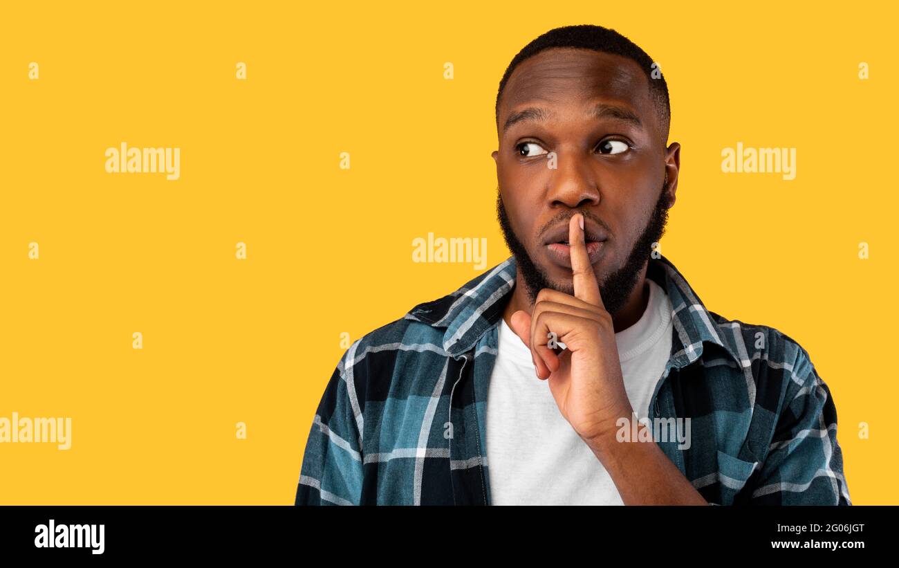 African american guy gesturing finger-on-lips looking aside over yellow ...