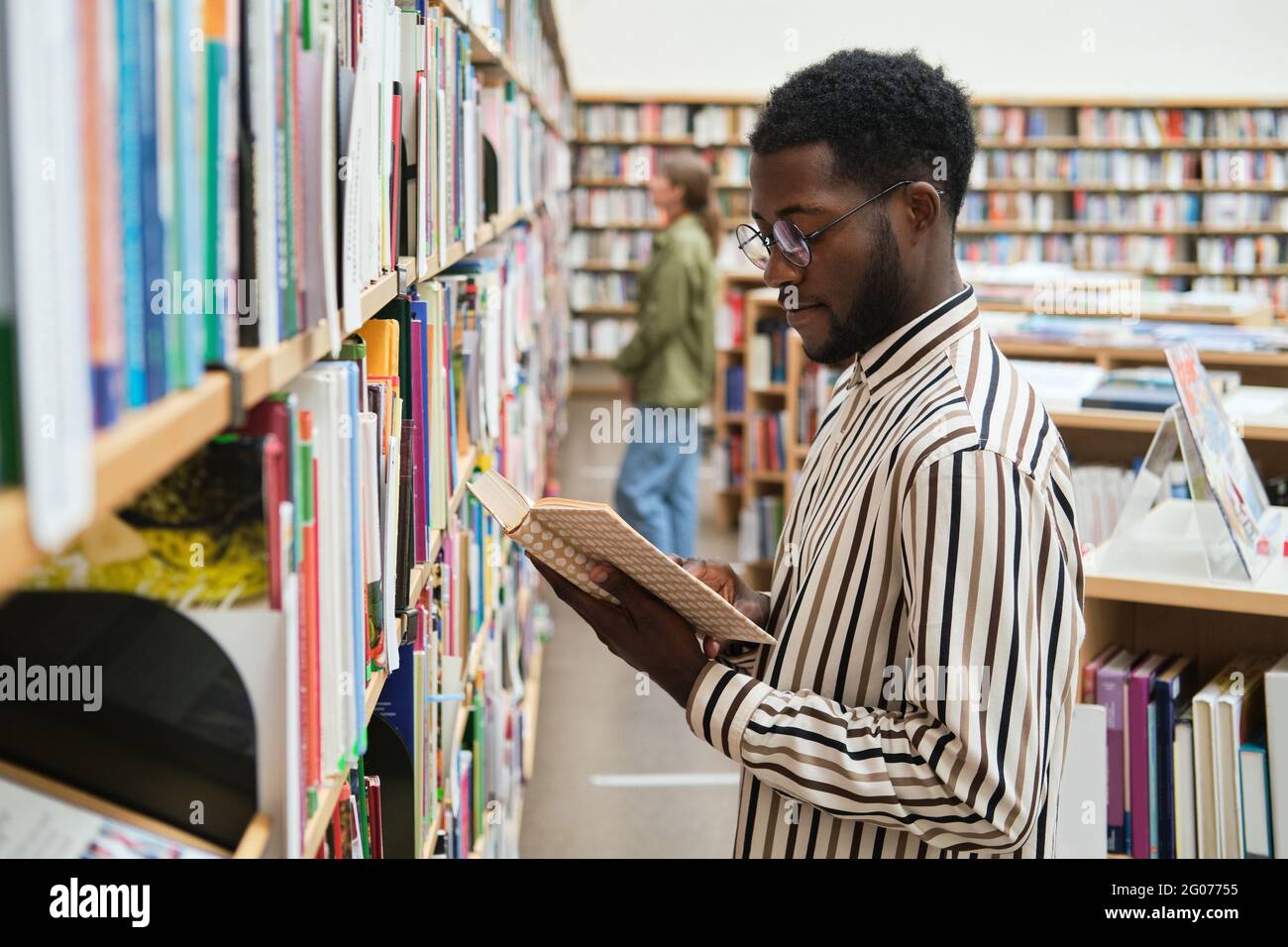 African student standing in front of the shelves and reading a book in ...