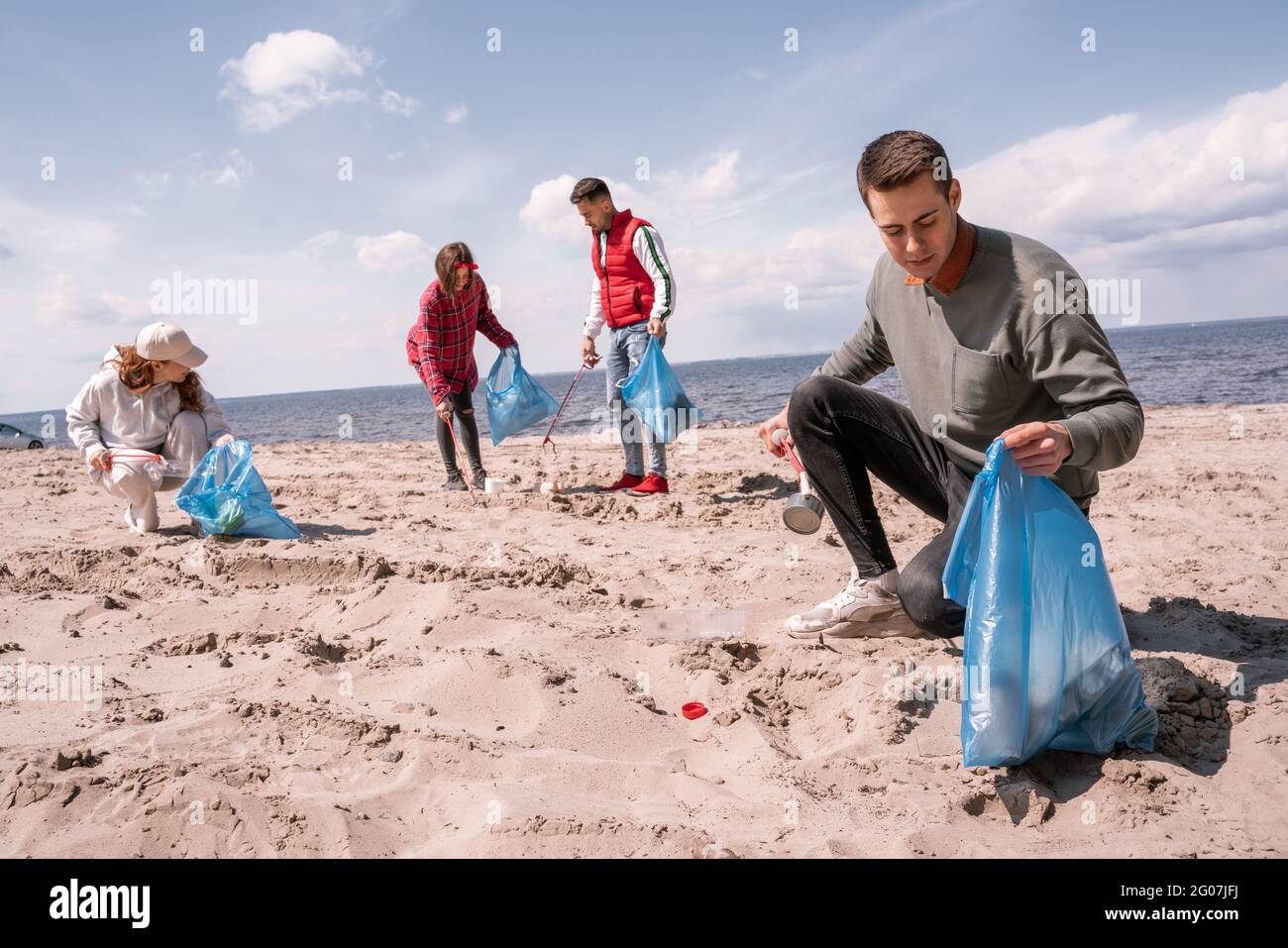 young man holding trash bag and collecting rubbish on sand near group ...