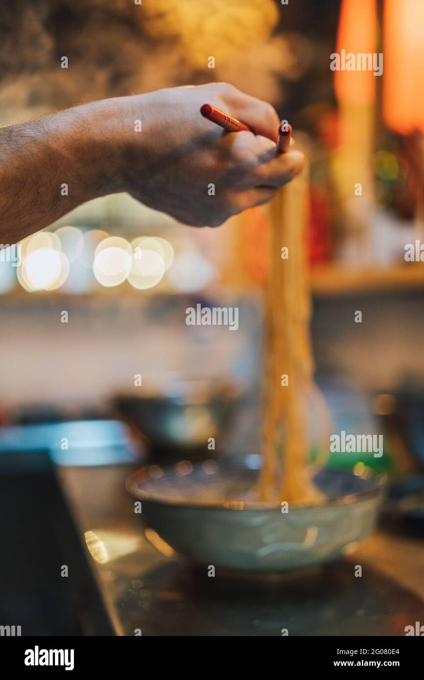 Crop hand of faceless chef with wooden chopsticks holding noodle above ...