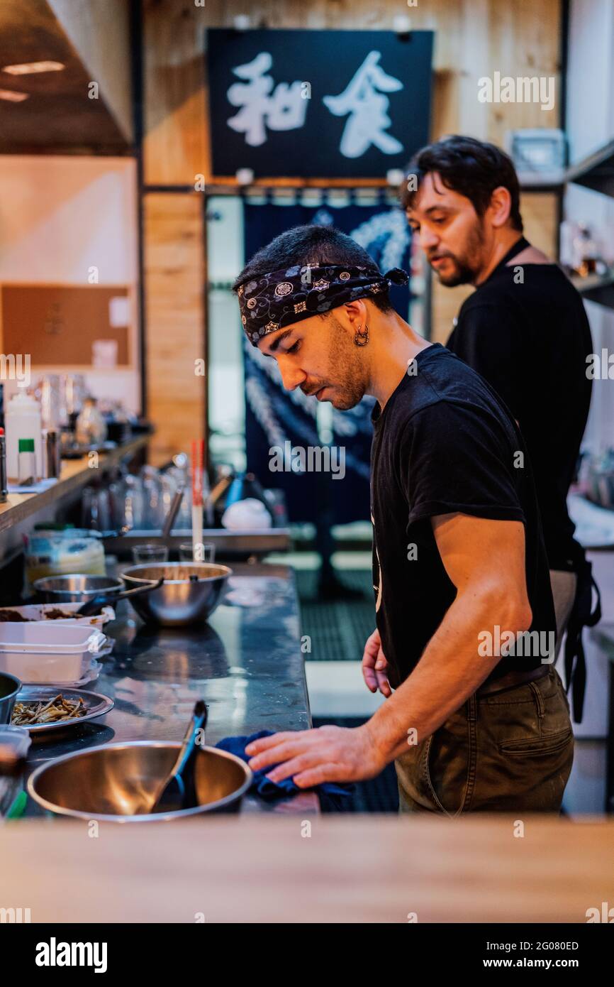 Side view of male chef in black uniform and bandana cooking Asian dish ...