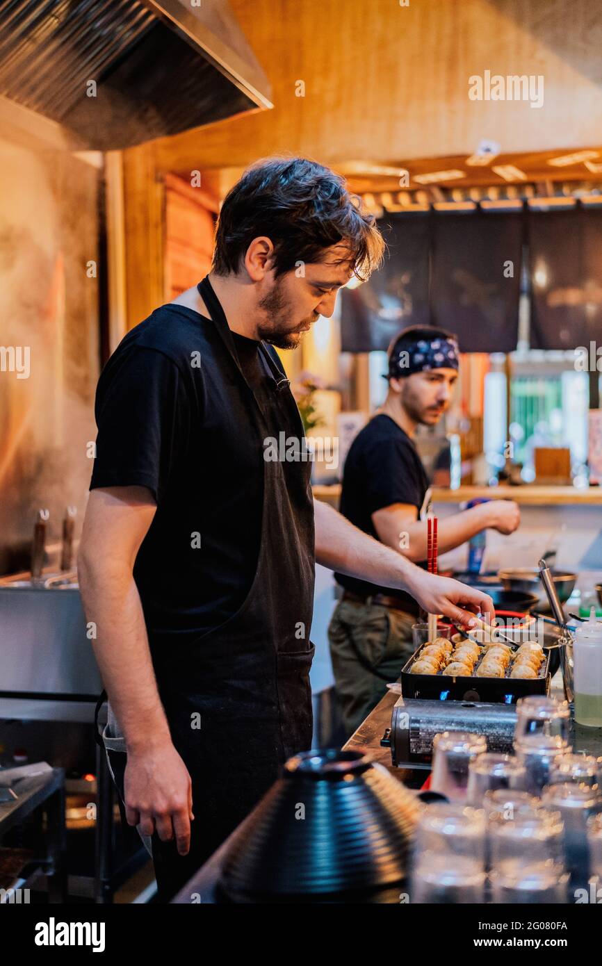 Side view of male chef in black uniform and bandana cooking Asian dish ...