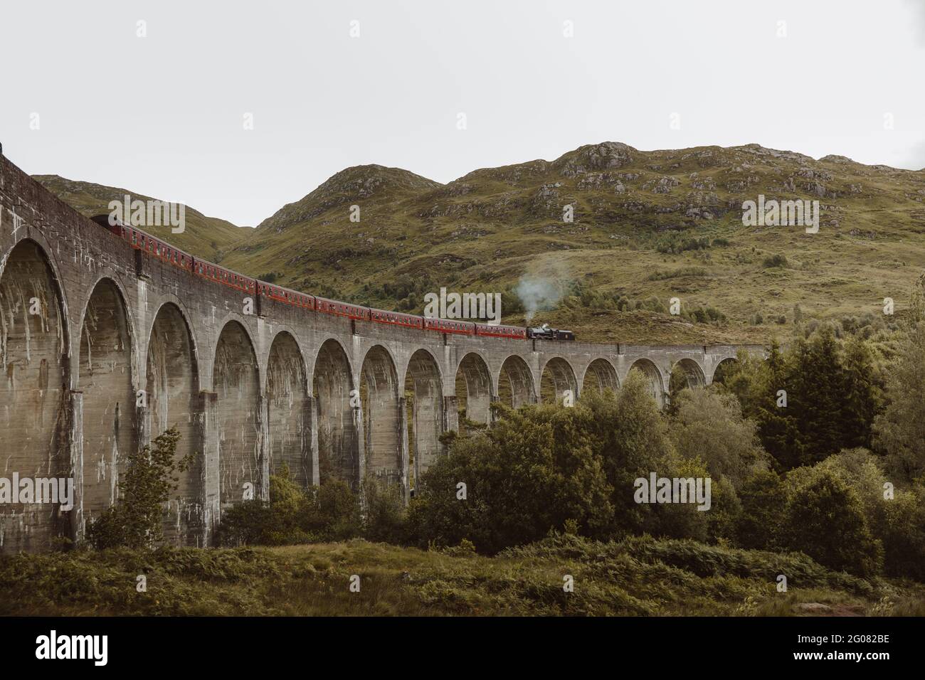 Steam train riding along old arch bridge near rough hill on gray day in ...