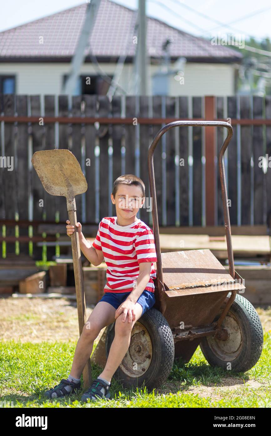 A little cheerful boy sits on a garden wheelbarrow and holds a shovel ...