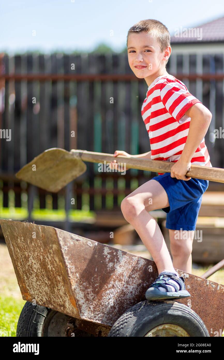 A little cheerful boy sits on a garden wheelbarrow and holds a shovel ...