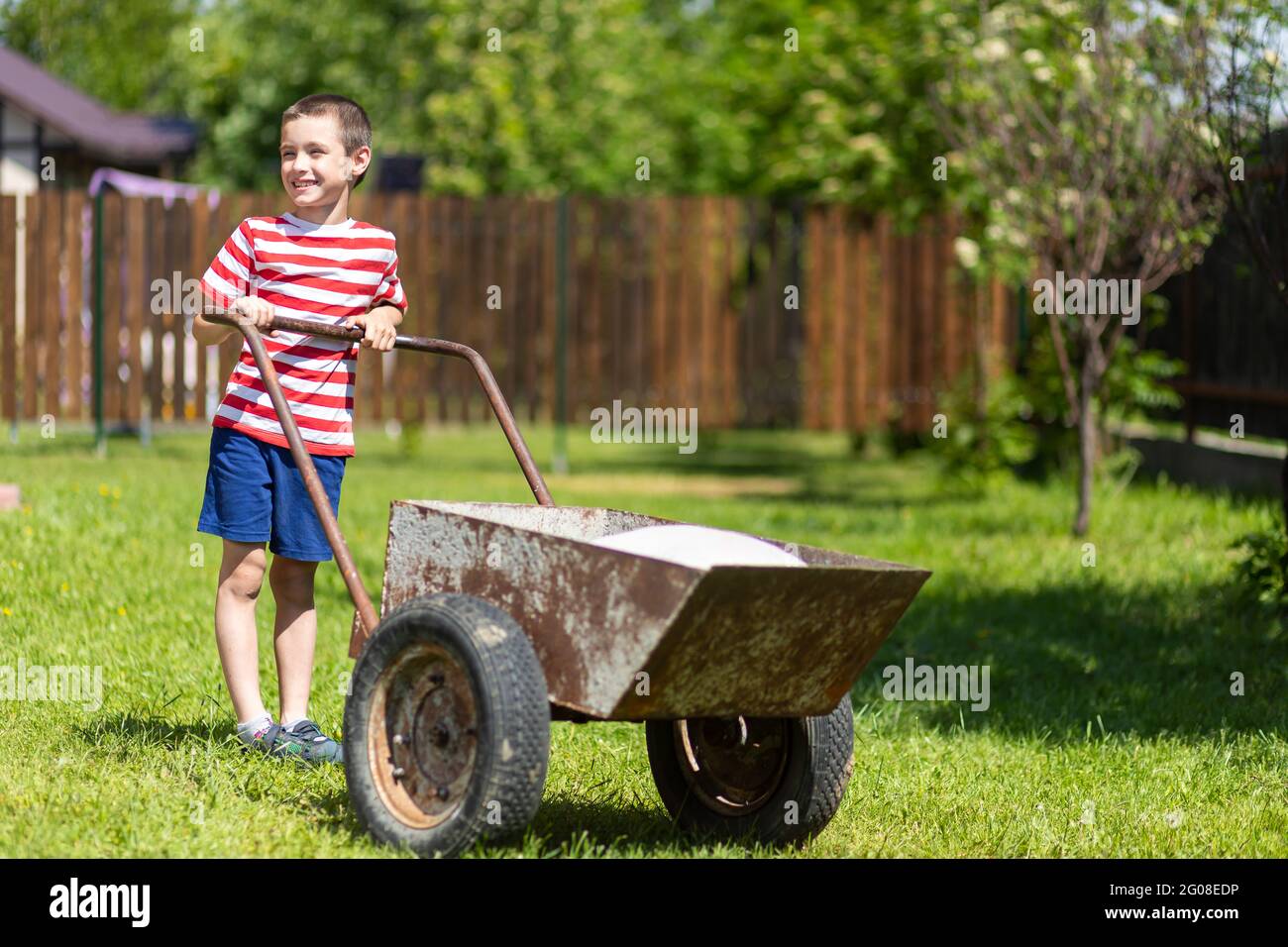 Young smile boy pushes a wheelbarrow around a yard.Boy helper in t ...