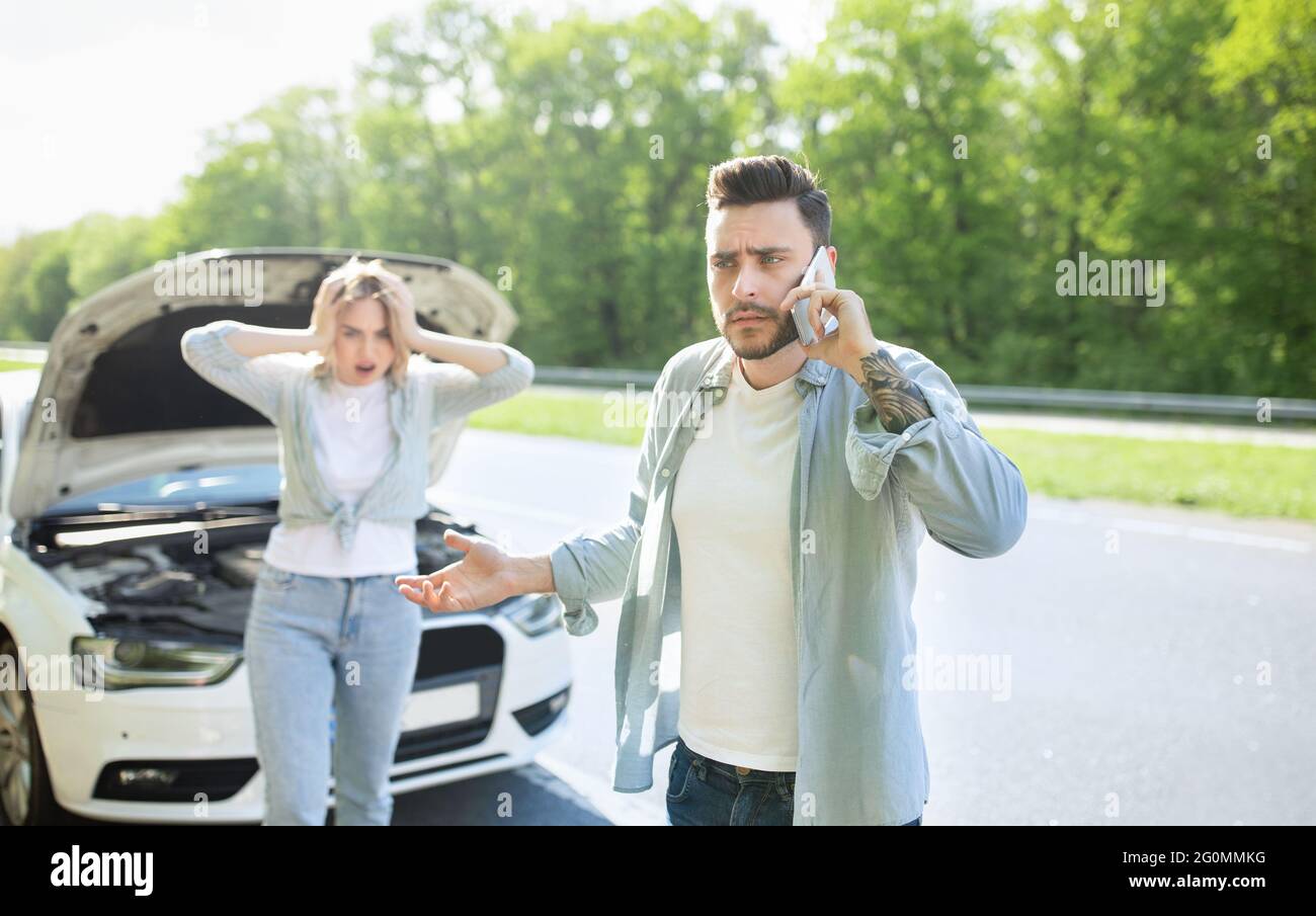Young guy standing near broken car together with terrified girlfriend ...