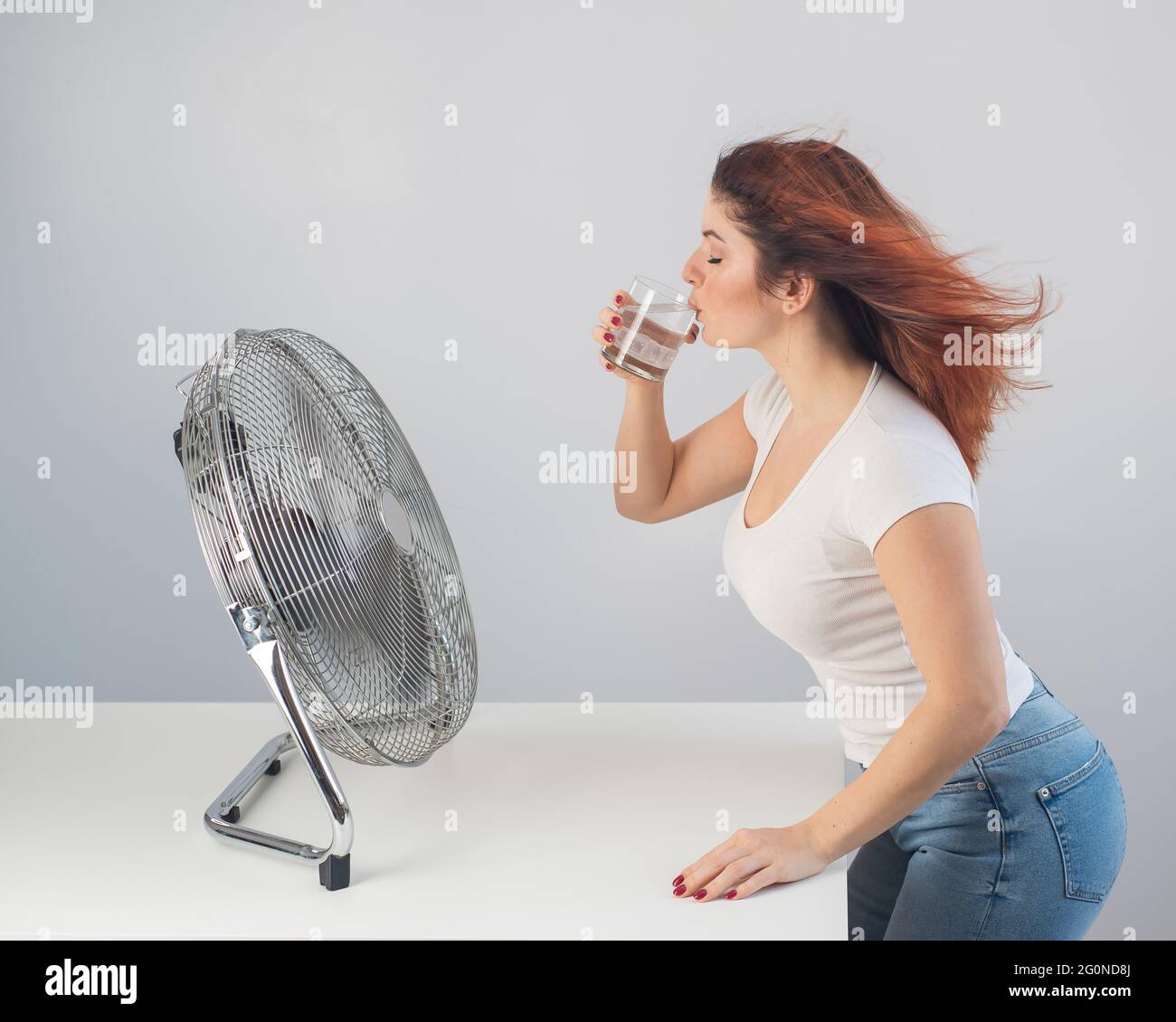 A red-haired Caucasian woman chills by the electric fan and drinks a ...