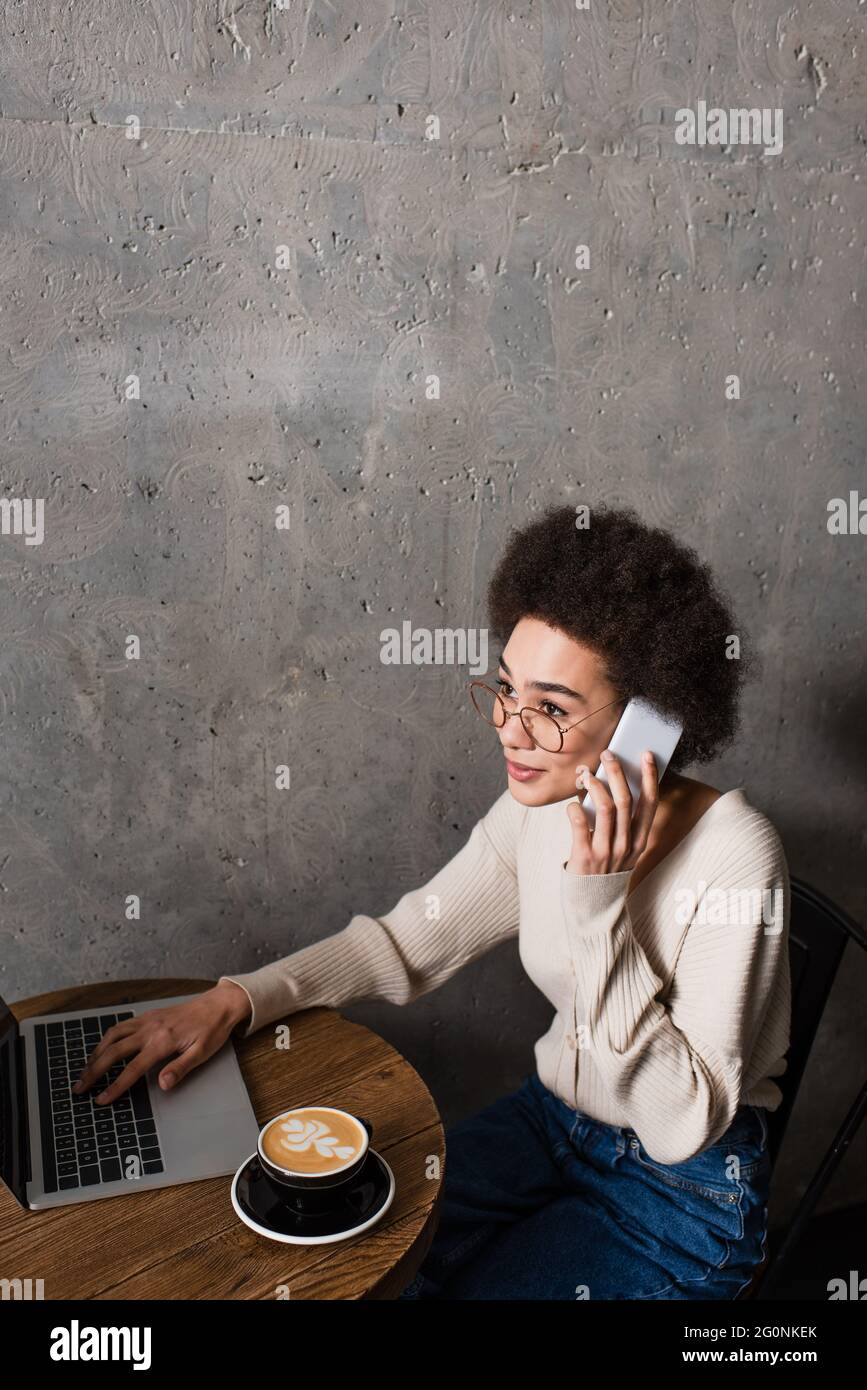 African american teleworker talking on smartphone and using laptop near ...