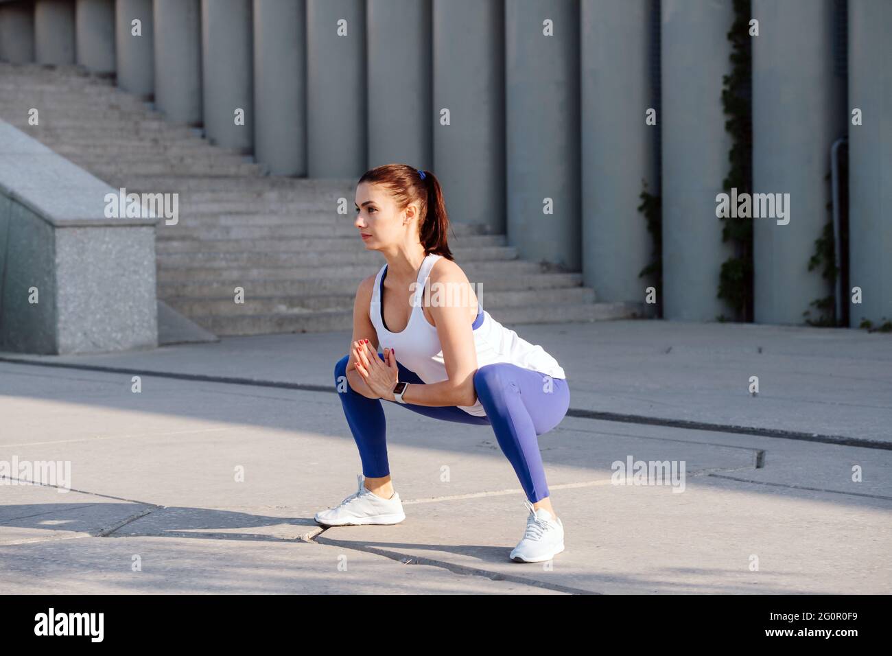 Resolved woman working out on the street, doing squat, hands joined ...