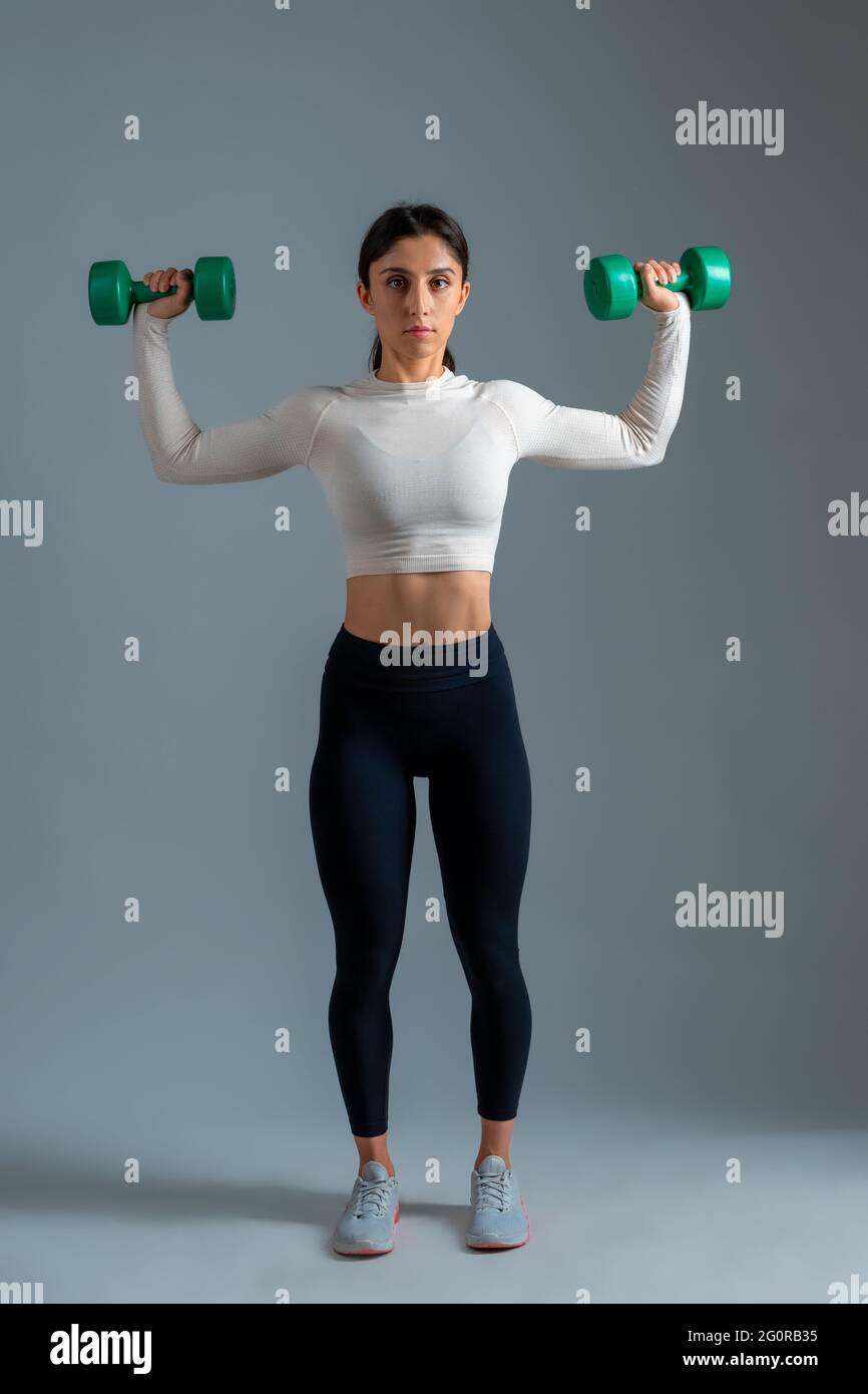 Young sportswoman doing dumbbell shoulder press on grey background ...