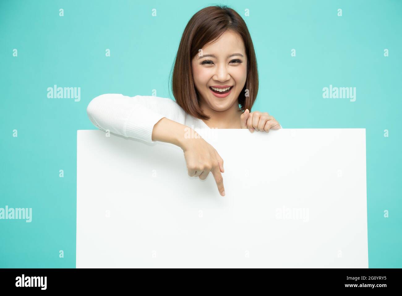 Smiling happy Asian woman standing behind big white poster and pointing finger down to blank ...
