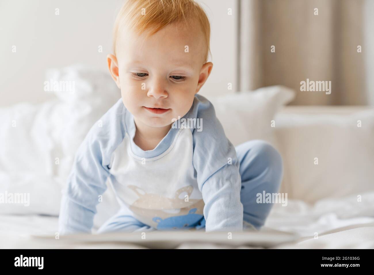 Focused blonde baby boy looking at tablet computer while playing on bed ...