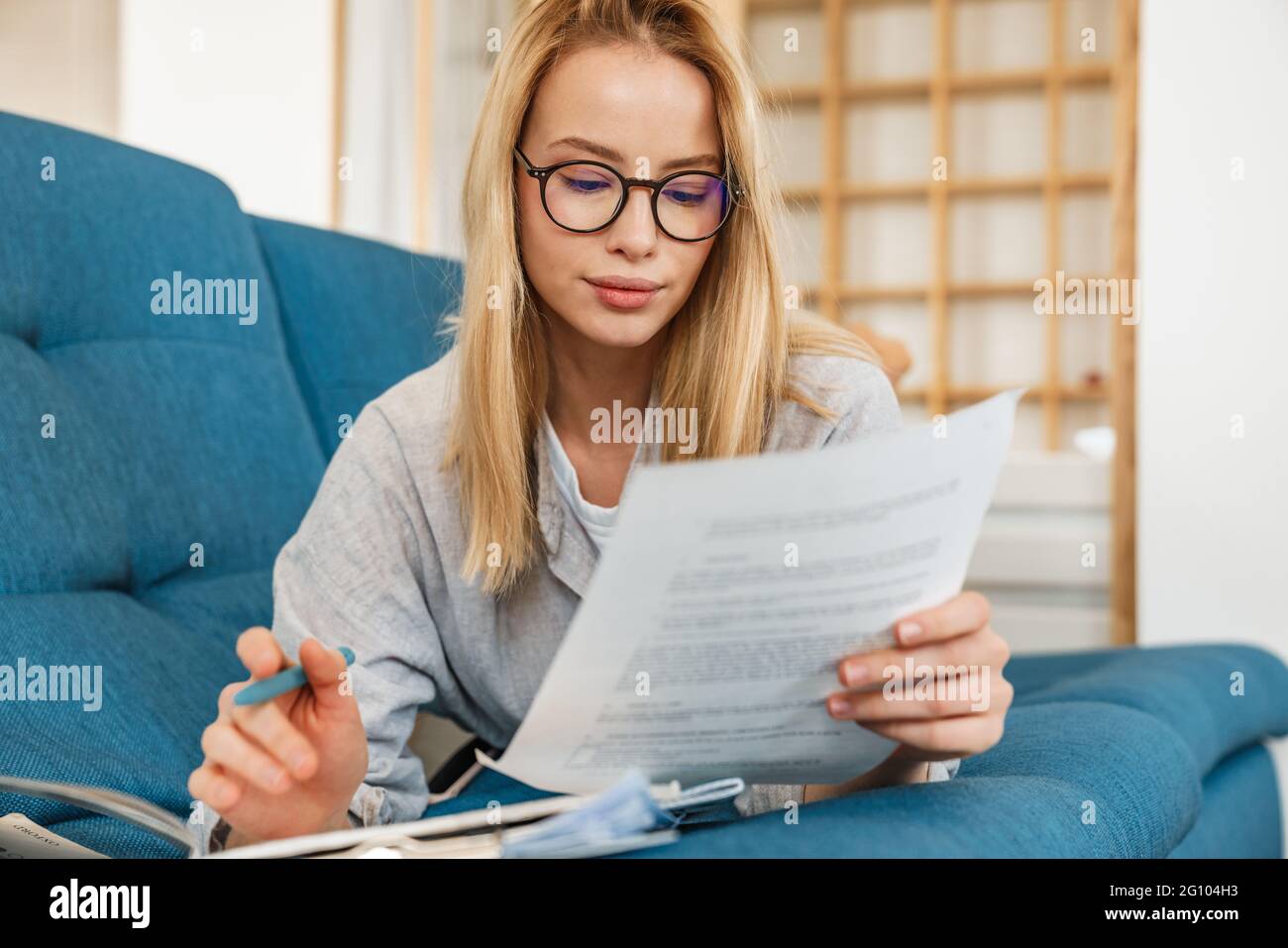 Focused student girl in eyeglasses doing homework while lying on couch ...
