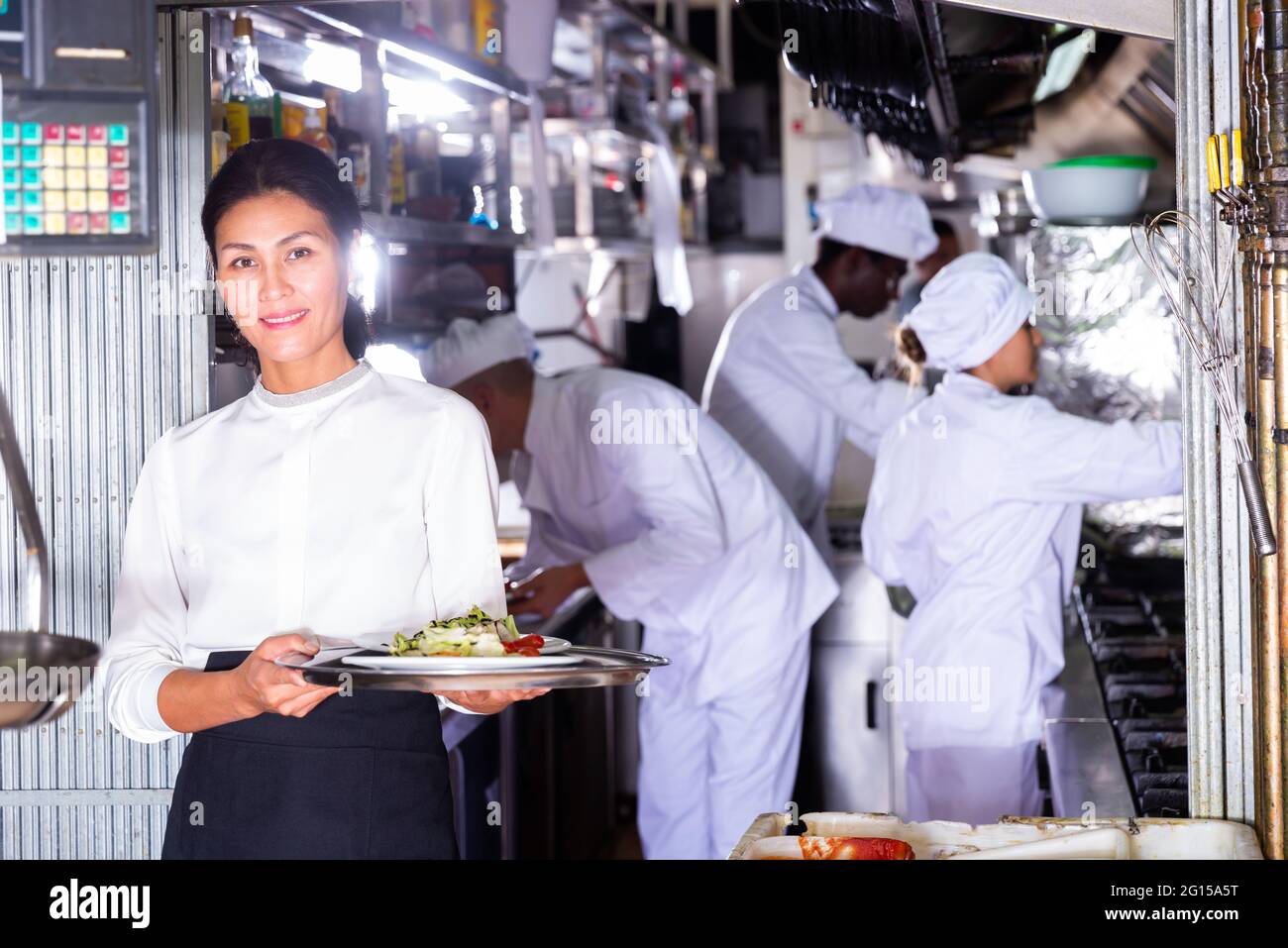 portrait of happy female waitress standing in white kitchen in cafe ...