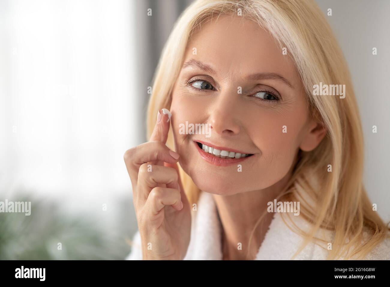 A woman in a white bath robe applying facial cream on her cheeks Stock ...