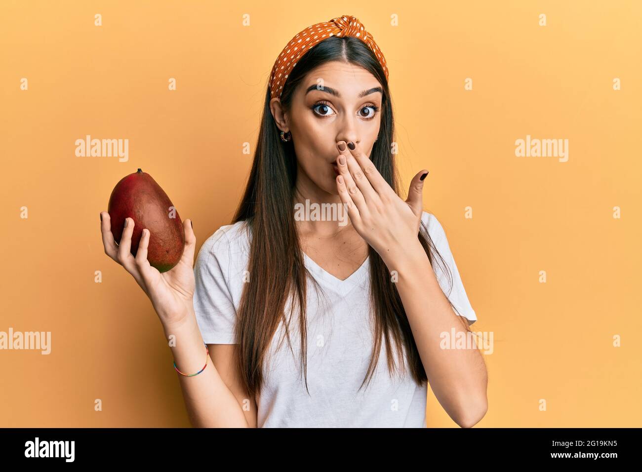 Beautiful brunette young woman holding mango covering mouth with hand ...