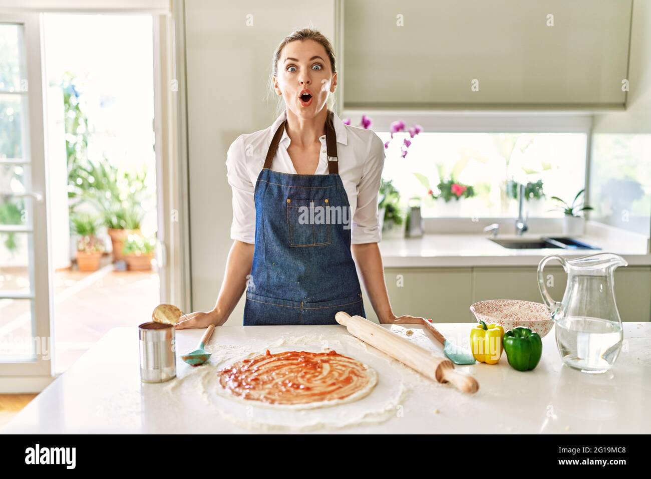 Beautiful blonde woman wearing apron cooking pizza scared and amazed ...