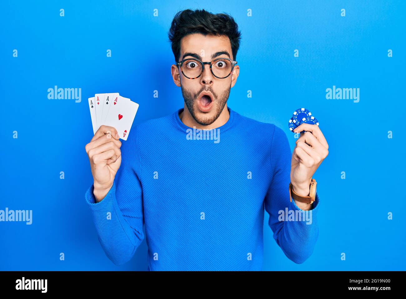 Young hispanic man playing poker holding casino chips and cards afraid ...
