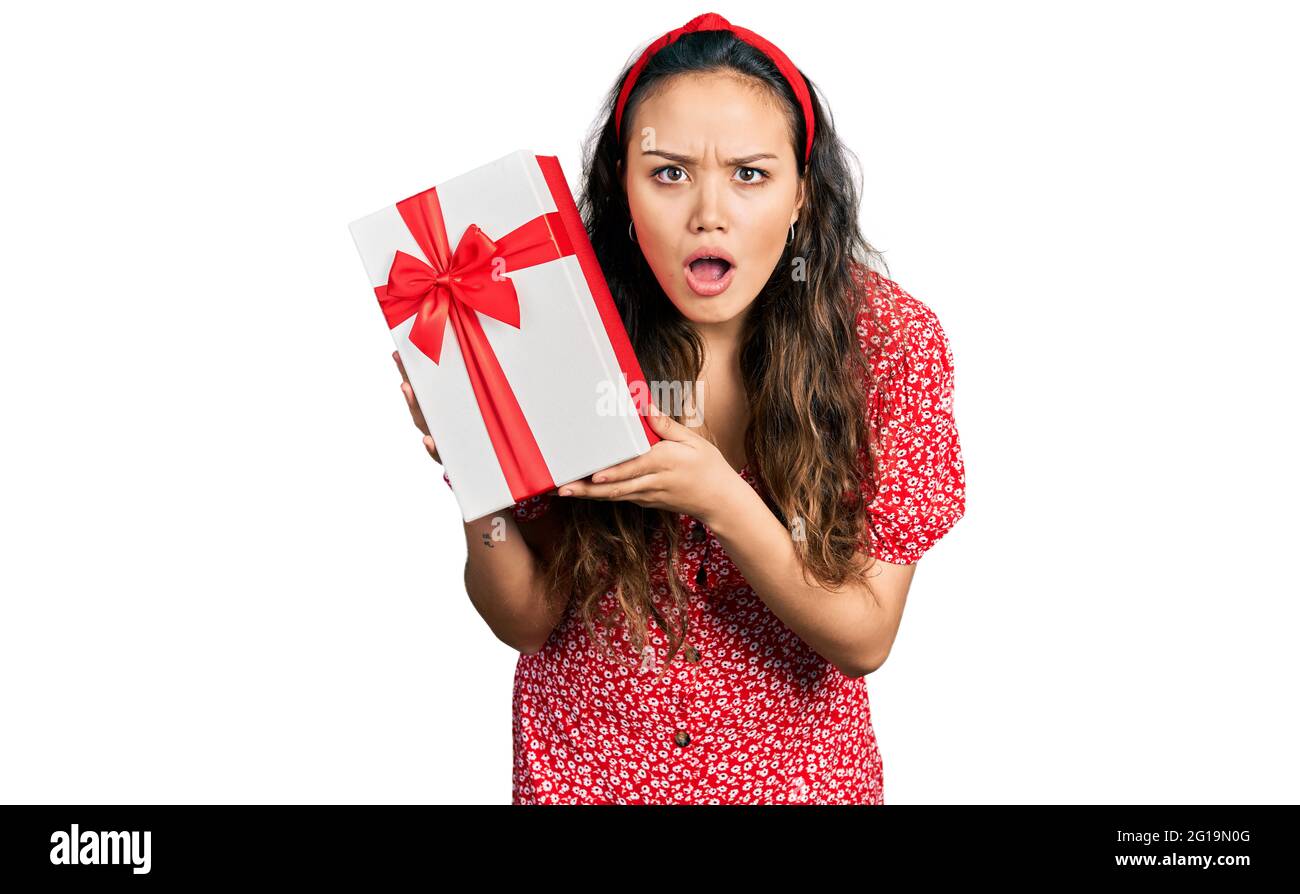 Young hispanic girl holding gift in shock face, looking skeptical and ...