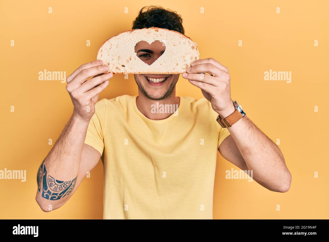 Young hispanic man holding bread with heart shape over eye smiling and ...