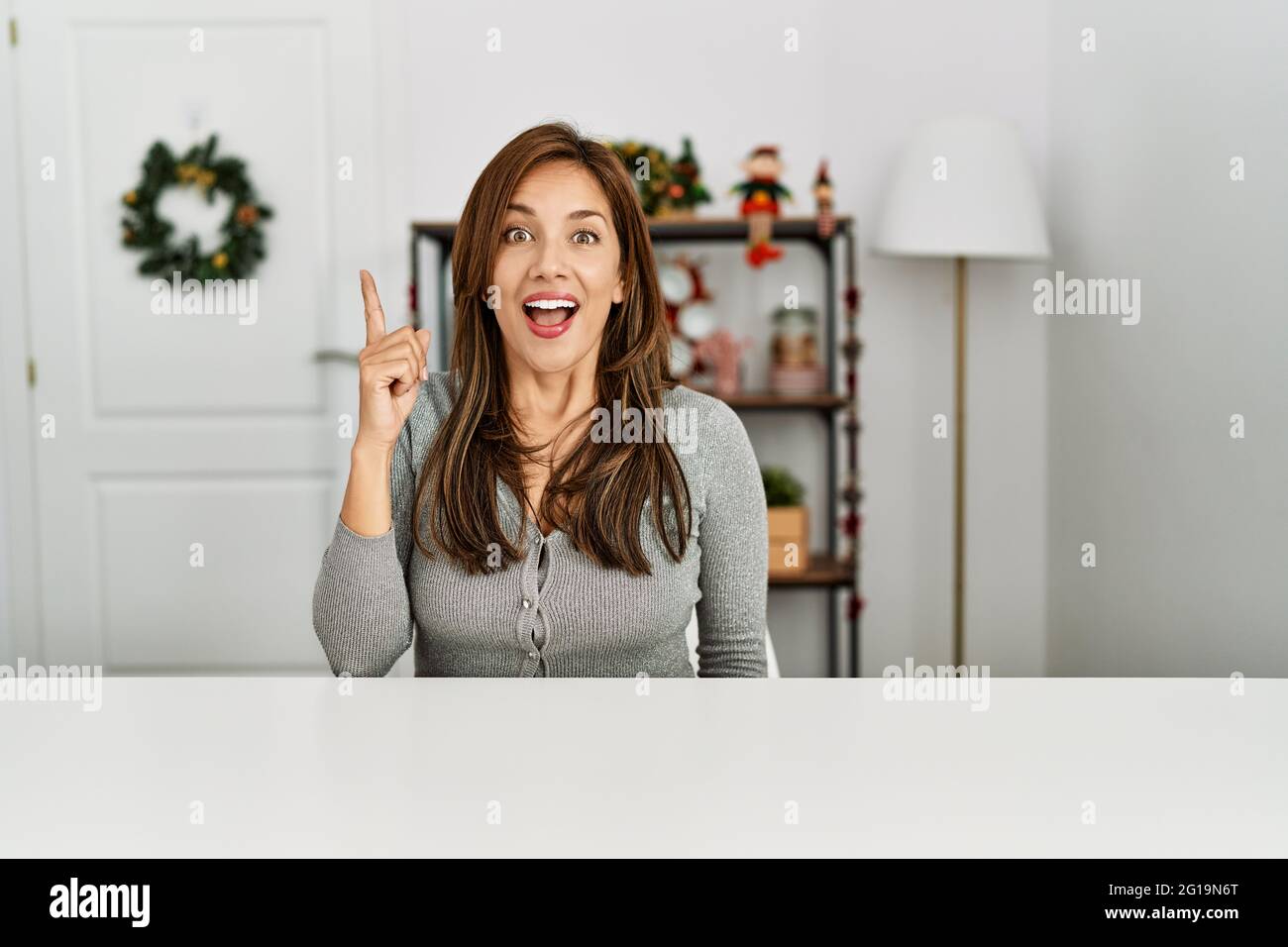 Young latin woman sitting on the table by christmas decor pointing ...