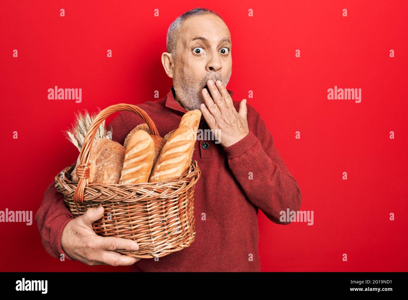 Handsome mature man holding wicker basket with bread covering mouth ...