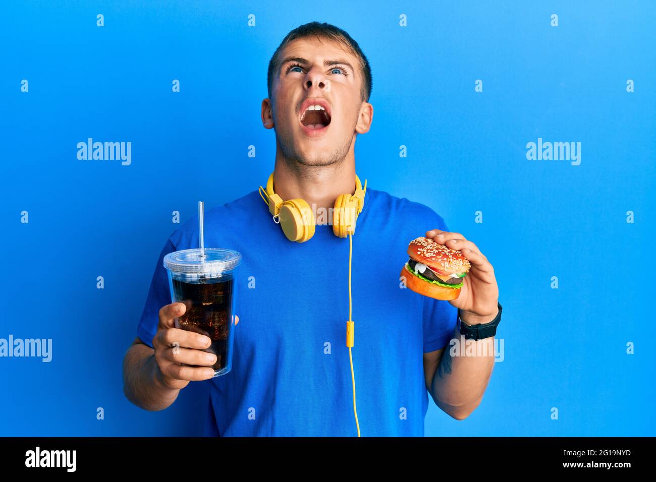 Young caucasian man eating a tasty classic burger and soda angry and ...