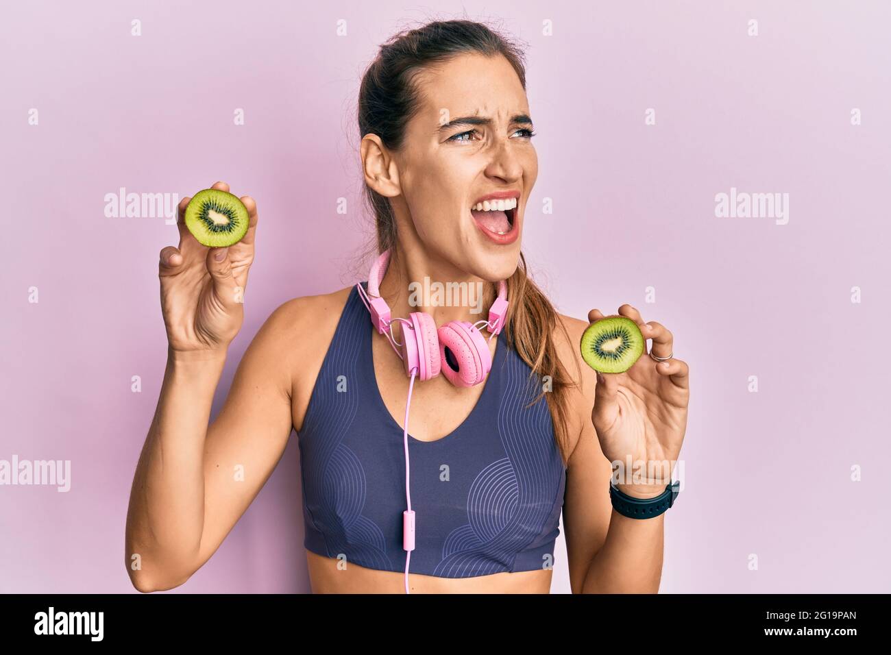 Young beautiful woman wearing gym clothes holding slice of kiwi angry ...