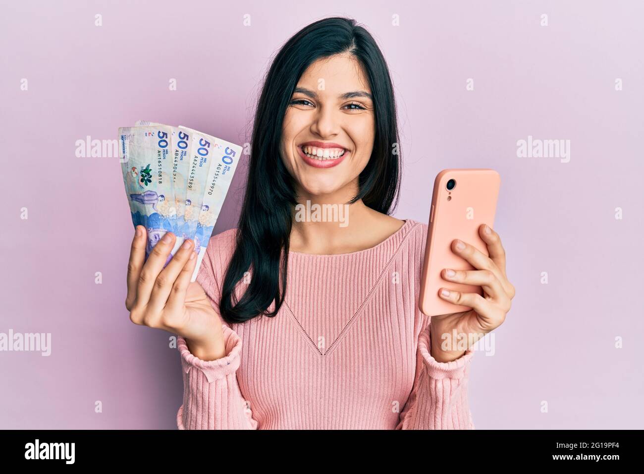 Young hispanic woman using smartphone holding colombian pesos banknotes ...