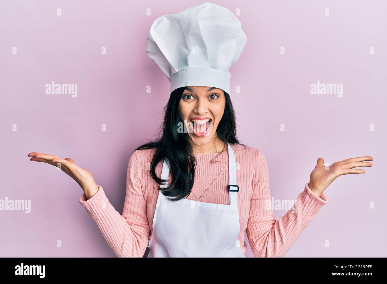 Young hispanic woman wearing baker uniform and cook hat celebrating victory with happy smile and ...