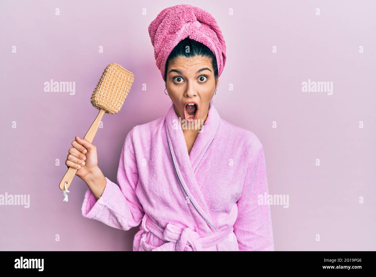 Young hispanic woman wearing shower bathrobe holding sponge scared and ...