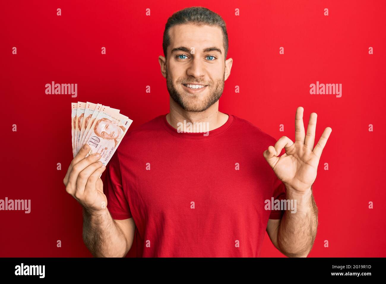 Young caucasian man holding 10 singapore dollars banknotes doing ok ...