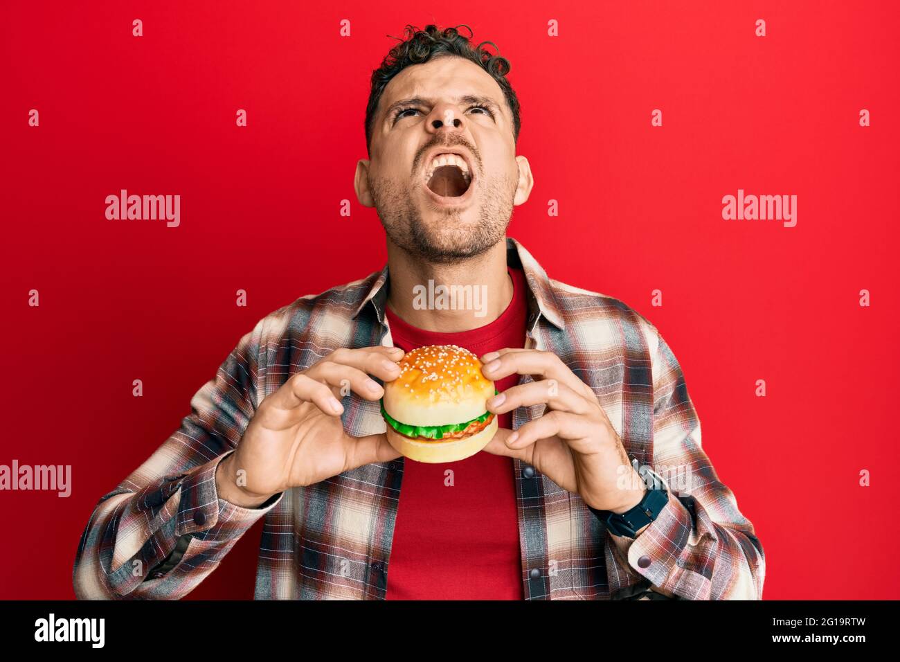 Young hispanic man eating a tasty classic burger angry and mad ...