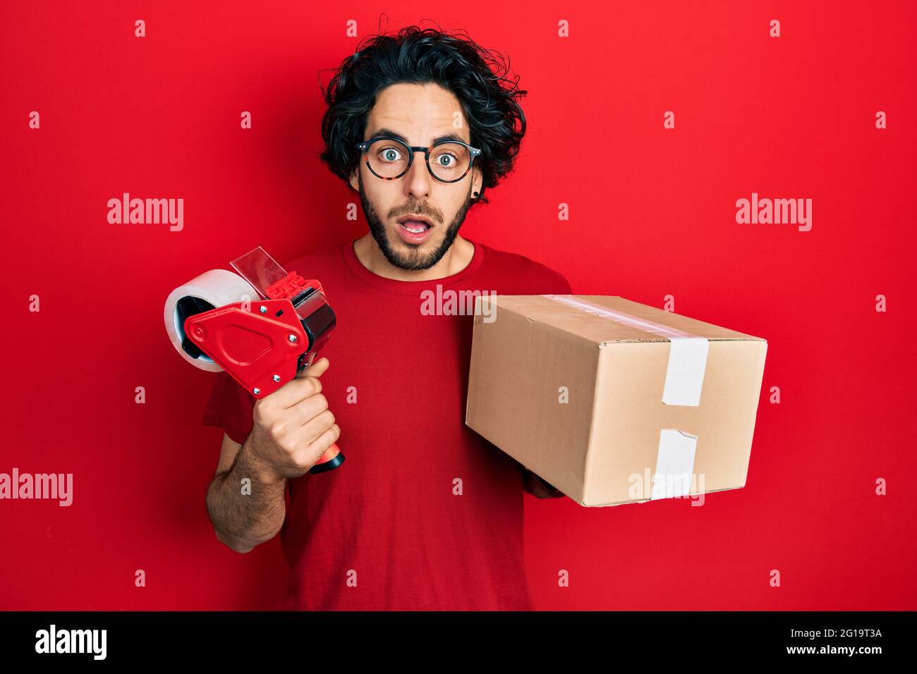 Handsome hispanic man holding packing tape and cardboard box afraid and ...