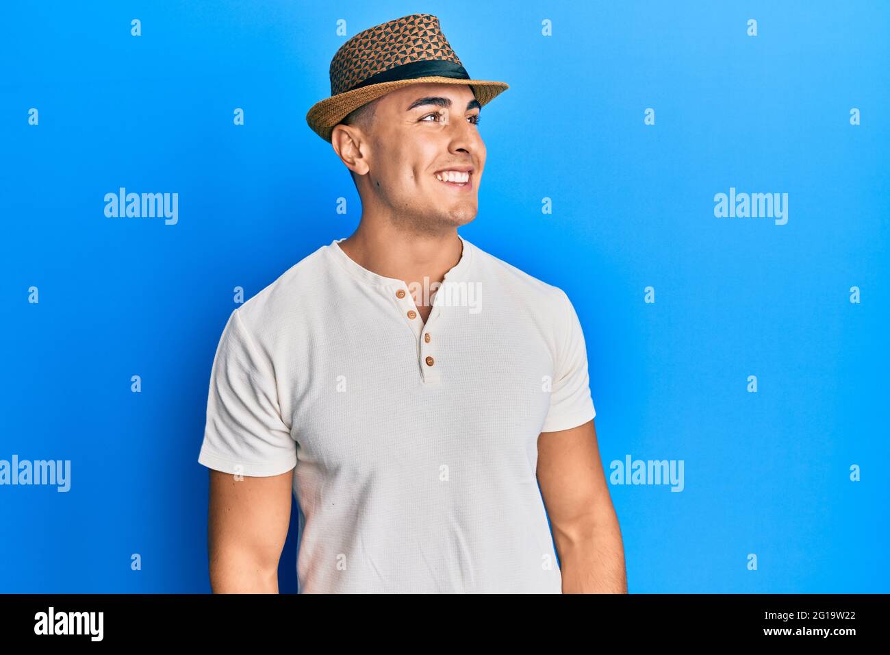 Hispanic young man wearing summer hat looking away to side with smile ...