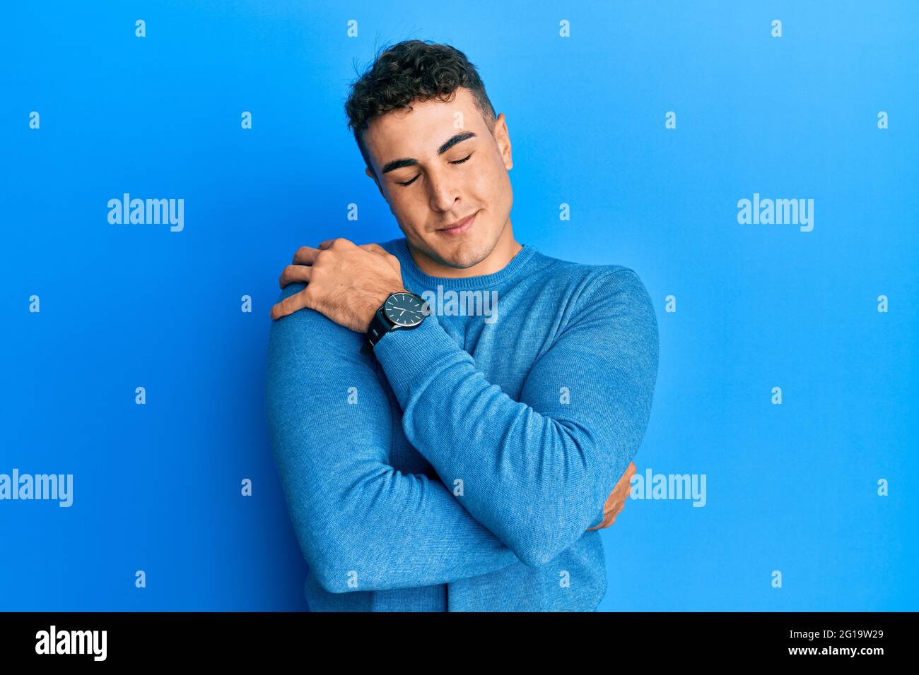 Hispanic young man wearing casual winter sweater hugging oneself happy ...