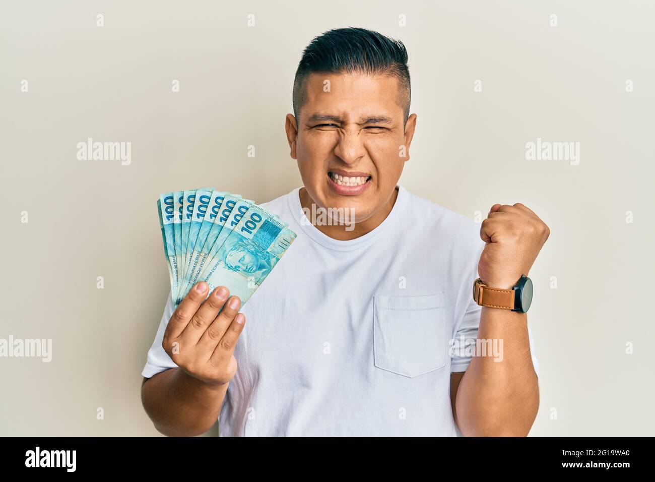 Young latin man holding 100 brazilian real banknotes screaming proud ...