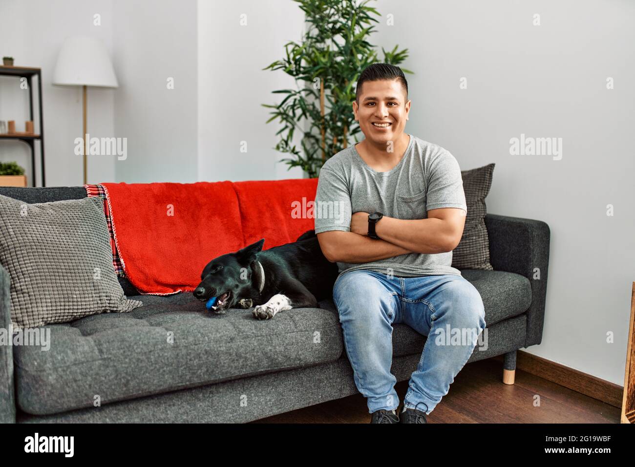 Young latin man and dog sitting on the sofa at home happy face smiling ...