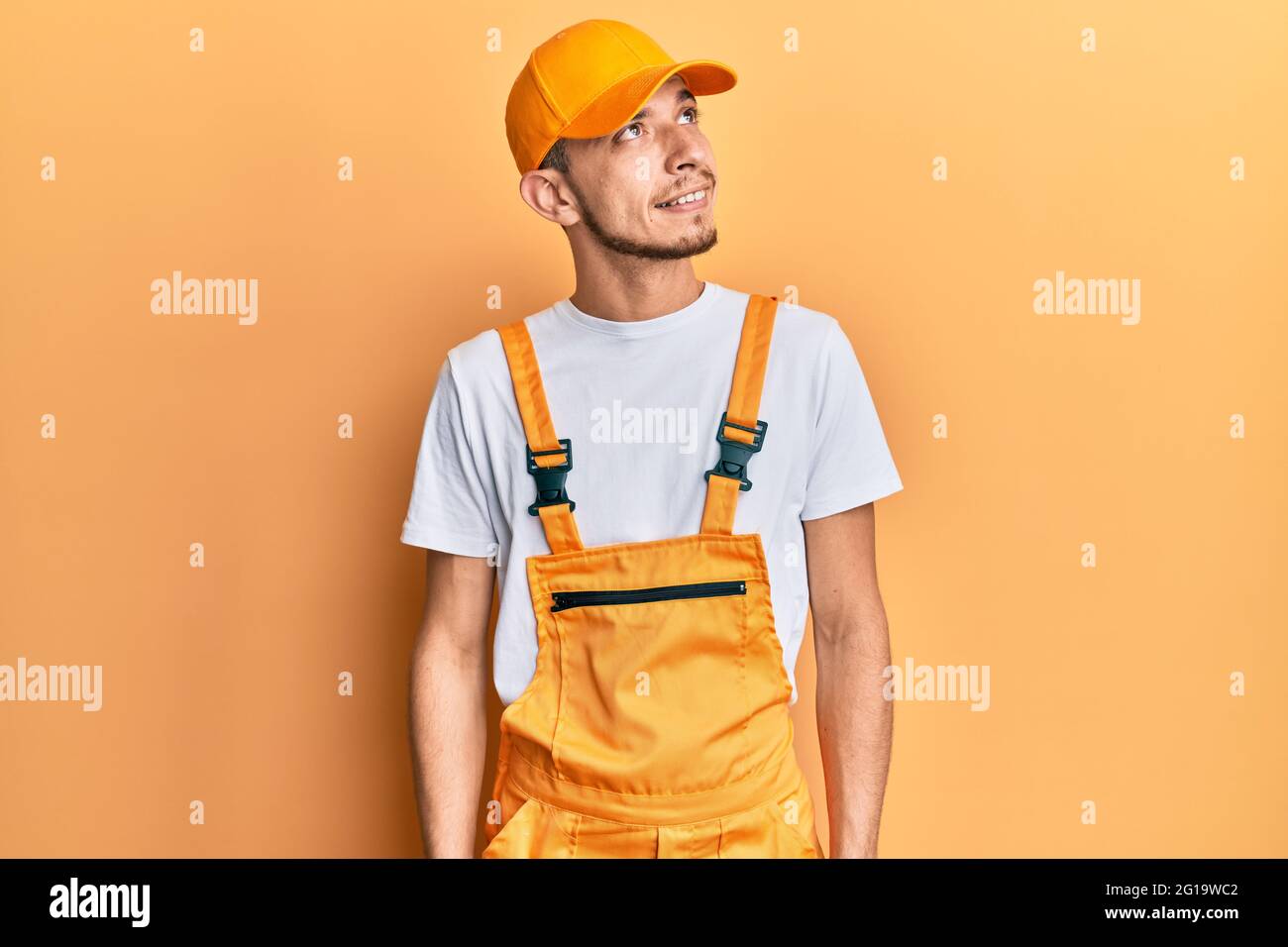 Hispanic young man wearing handyman uniform looking away to side with ...