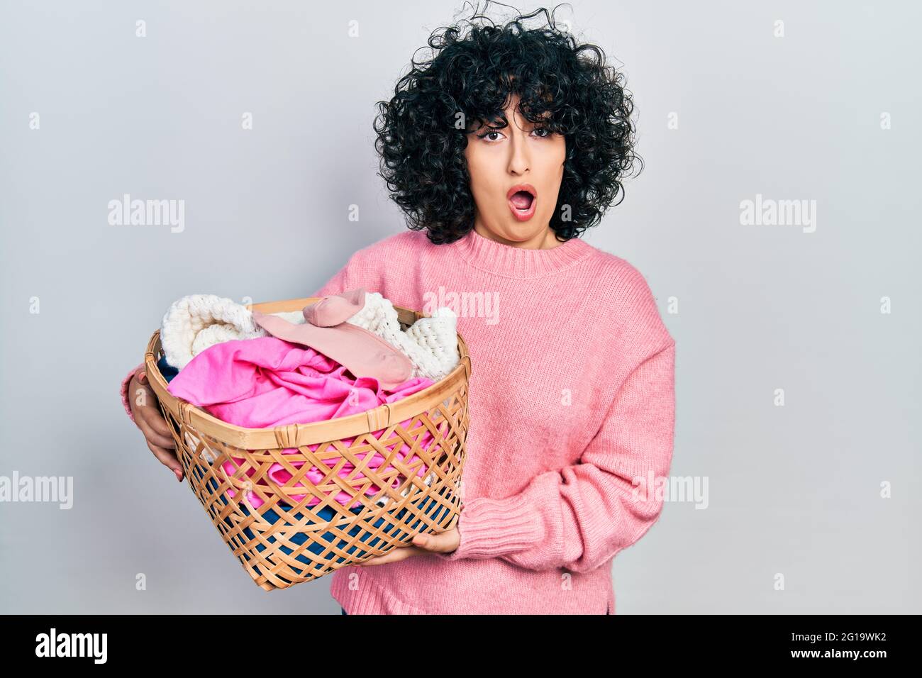 Young middle east woman holding laundry basket in shock face, looking ...