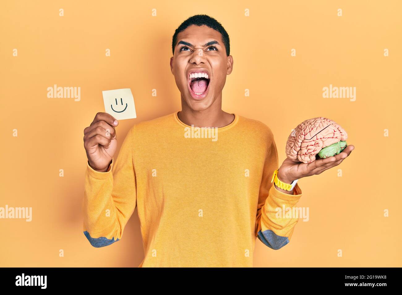 Young african american guy holding smile reminder and brain angry and ...