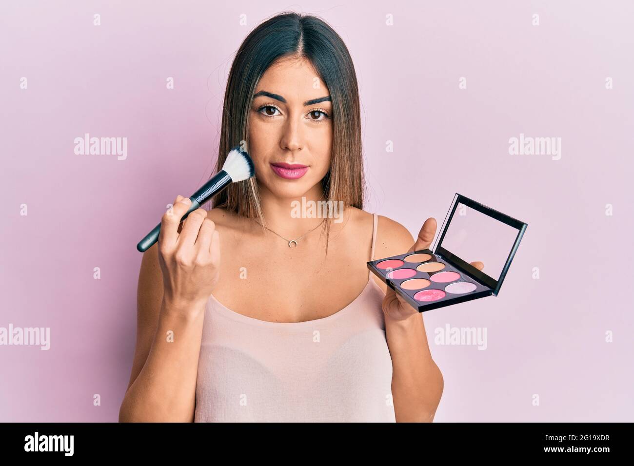 Young hispanic woman holding makeup brush and blush relaxed with ...
